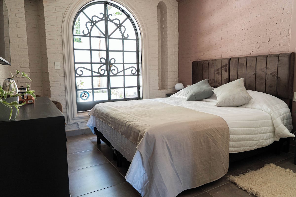 Quarto com uma cama grande, cabeceira de madeira, parede branca e janela com grade de ferro decorativa.