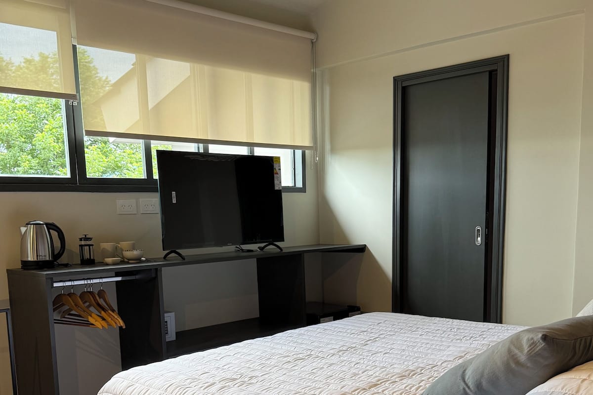Quarto com cama, escrivaninha com televisão, guarda-roupa e janela com persianas.