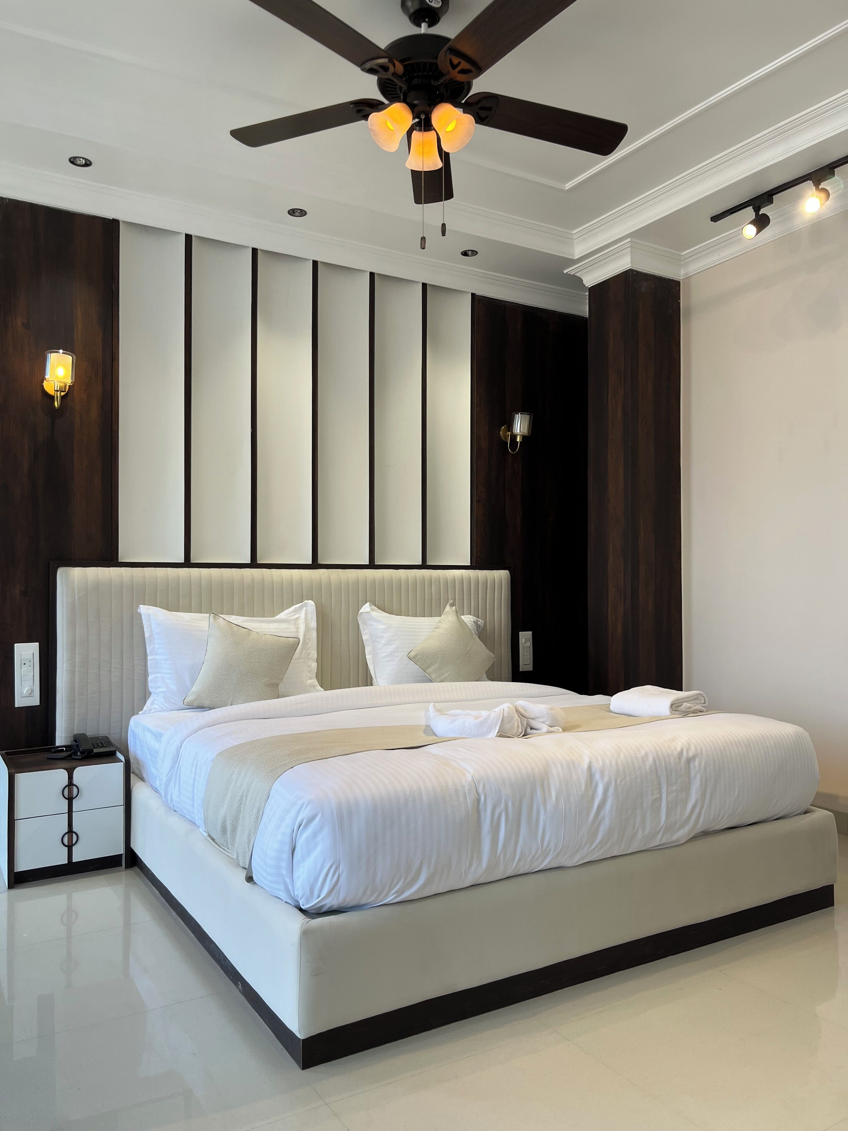 Quarto moderno com uma cama grande, ventilador de teto, iluminação embutida e mesa de cabeceira com telefone.