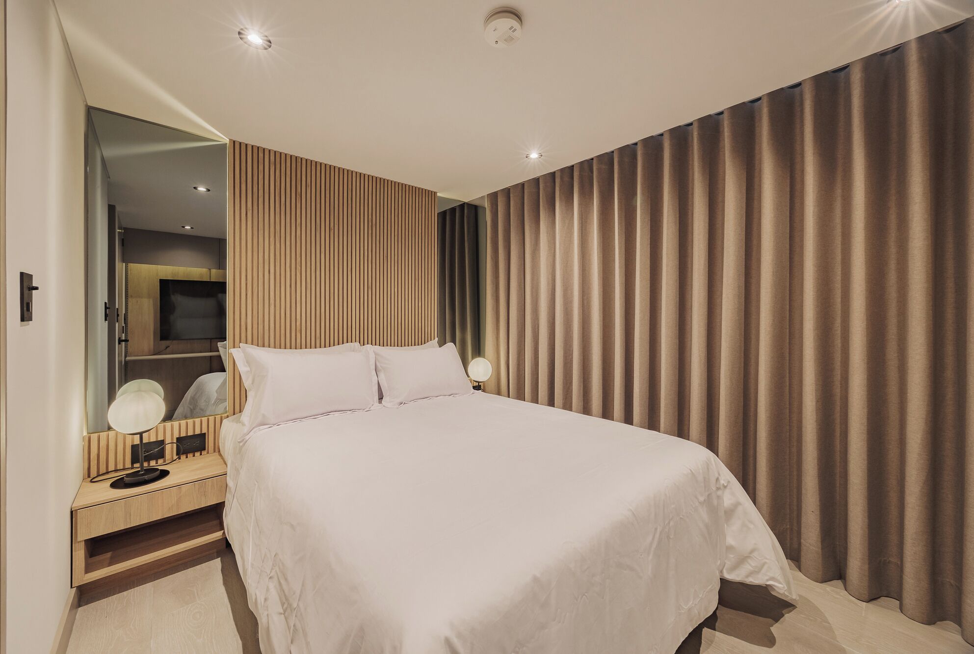 Quarto de hotel moderno com uma cama grande, televisão de tela plana, mesa de cabeceira com abajur e uma janela ampla com cortinas.