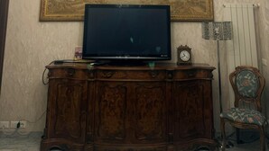 Quarto com lustre, televisão sobre um armário de madeira, uma pintura grande emoldurada e uma poltrona antiga.