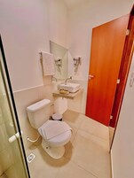 Casa de banho compacta com sanita, lavatório e espelho em cima. Há uma toalha pendurada na parede e uma porta à direita.