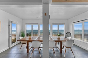 Uma área de refeições com vista para o oceano, mesa de madeira, cadeiras brancas e uma varanda com guarda-corpo.