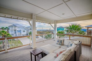 Uma área externa coberta com um deck de madeira, um sofá branco com almofadas e uma pequena mesa.