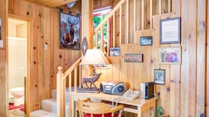 Interior de madeira com escada, escrivaninha com luminária, cadeira, telefone e quadros nas paredes.
