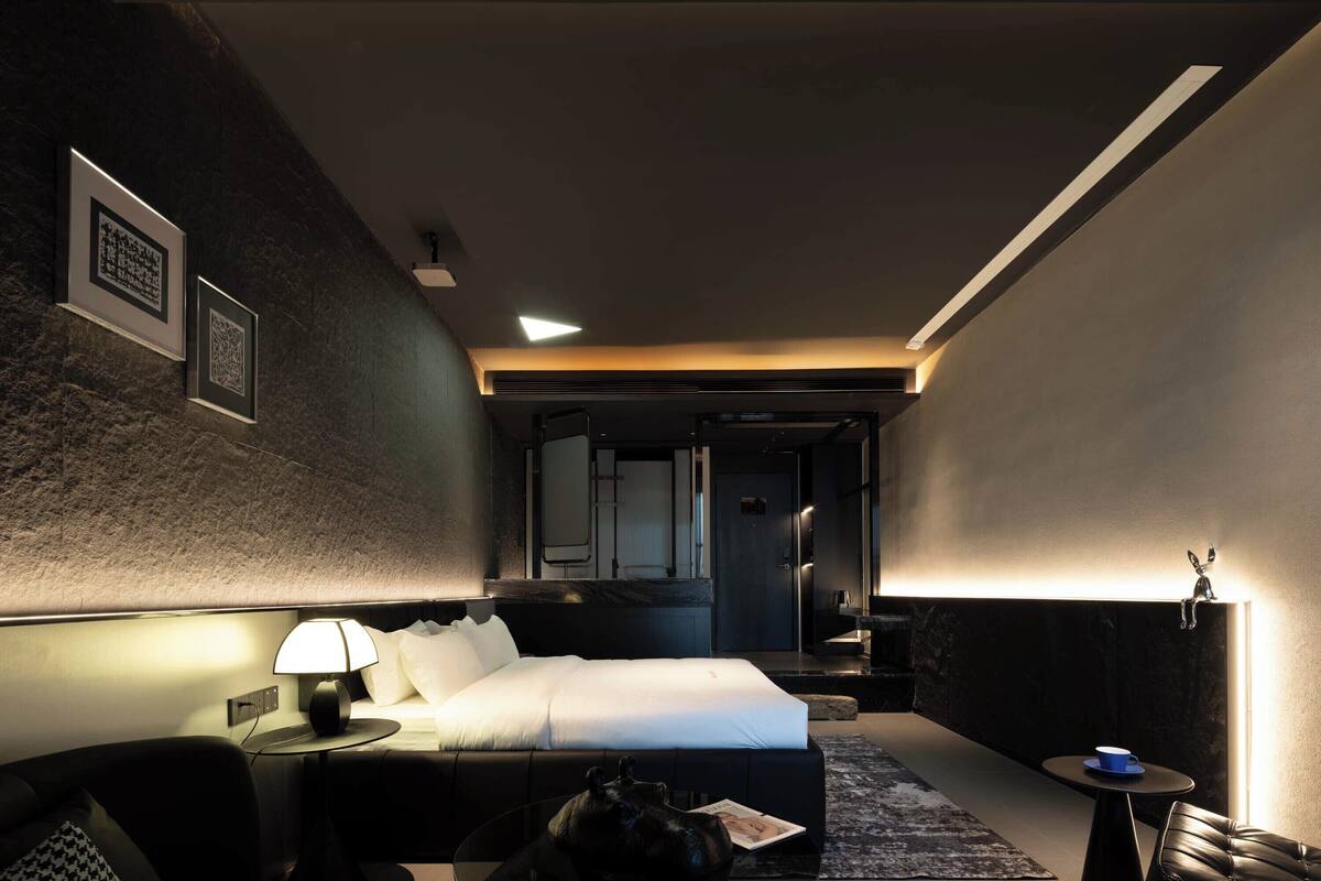 Quarto de hotel moderno com uma cama grande, mesas de cabeceira, uma escrivaninha e uma cadeira. Há uma TV fixada na parede e um quadro emoldurado.