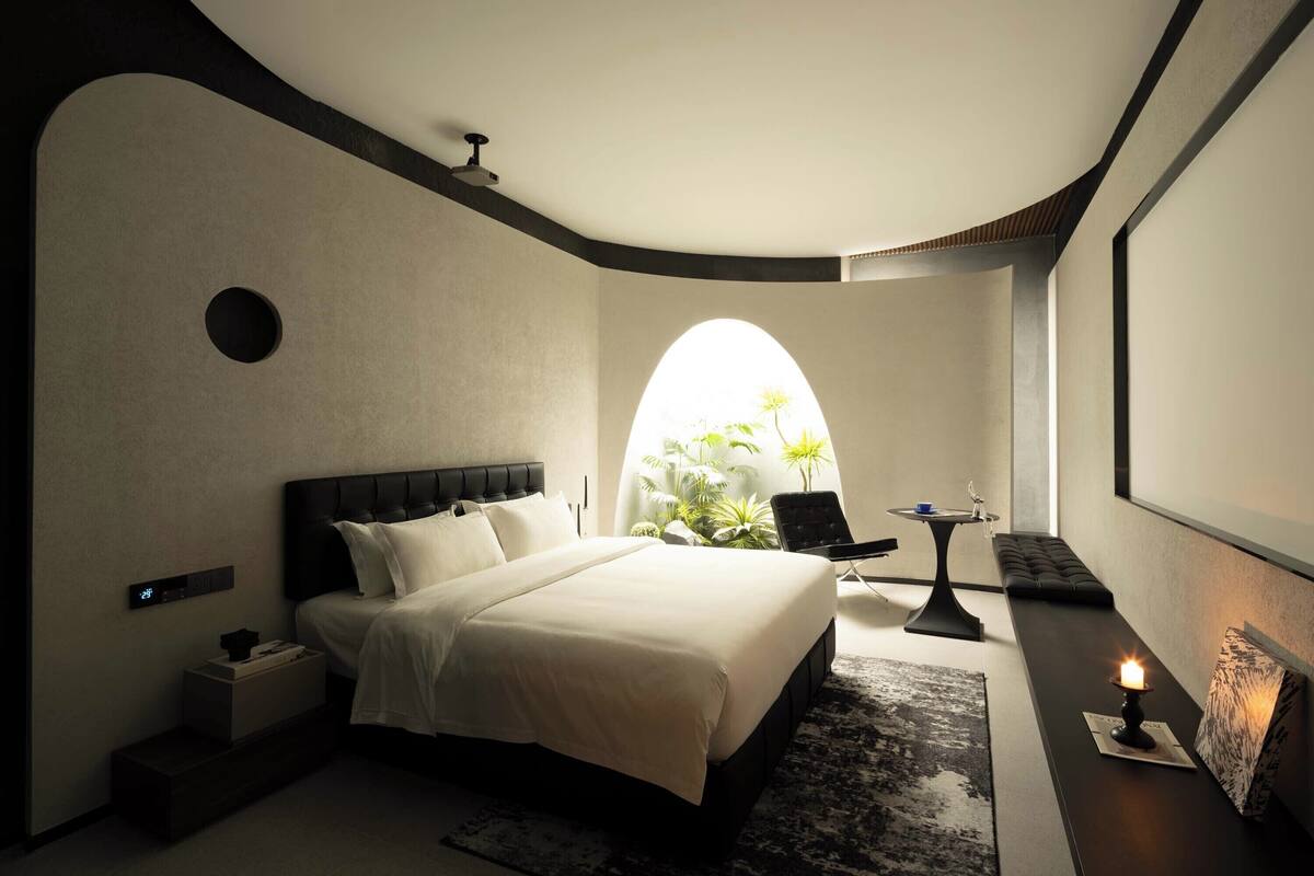 Quarto moderno com uma cama grande, mesas de cabeceira, uma pequena mesa e um espaço de estar com vista para vegetação.