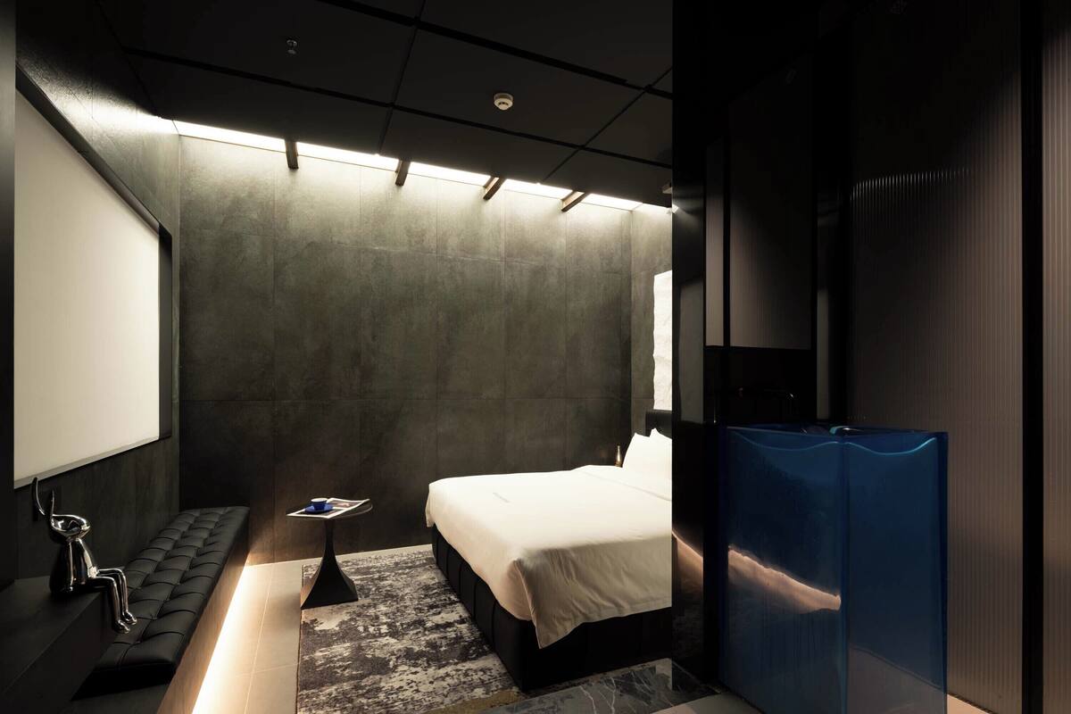 Quarto moderno com cama grande, mesinha de cabeceira, um banquinho e TV fixada na parede.
