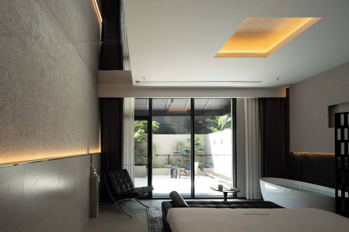 Quarto de hotel moderno com uma cama grande, área de estar com uma poltrona e mesa, e vista para o exterior por uma porta de vidro deslizante.