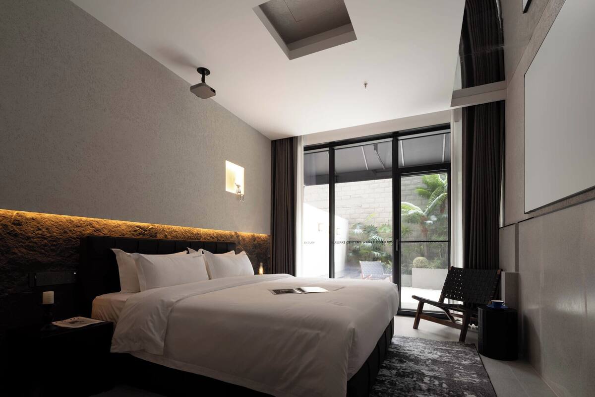 Quarto de hotel com cama grande, mesa de cabeceira, luminária, cadeira e vista para um pátio com plantas.