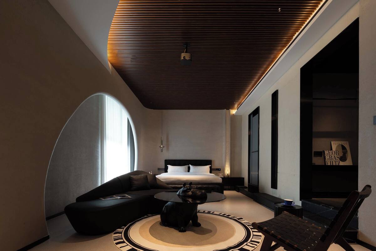 Quarto de hotel moderno com uma cama grande, um sofá preto, uma mesa de centro redonda e uma pequena estátua.