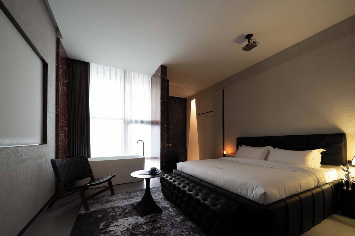 Quarto de hotel com uma cama grande, uma mesinha redonda pequena, uma cadeira e vista para o exterior pela janela.