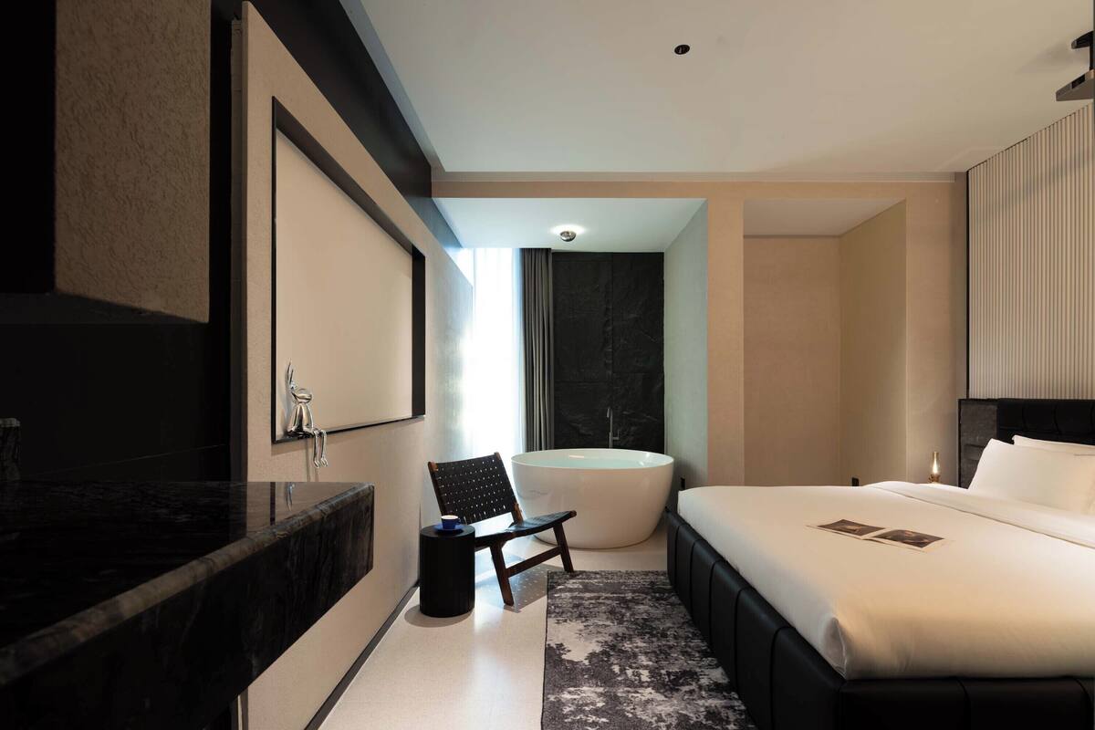 Quarto de hotel moderno com uma cama grande, banheiro separado com banheira, pequena área de estar e bancada de mármore.