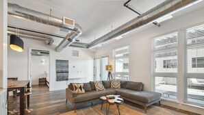 Uma sala de estar moderna com um sofá em formato de L, uma mesa de centro e um espaço de jantar com banquetas. Há grandes janelas com persianas e dutos visíveis no teto.