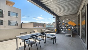 Um pátio externo com mesa e cadeiras, um espaço de estar acolchoado e um mural geométrico na parede.