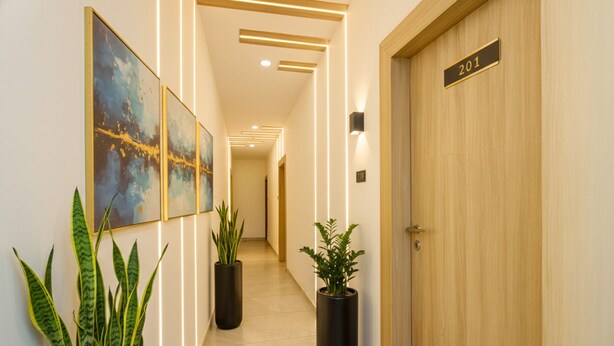 Corredor com porta de madeira com a placa “201”, plantas em vasos e quadros nas paredes.