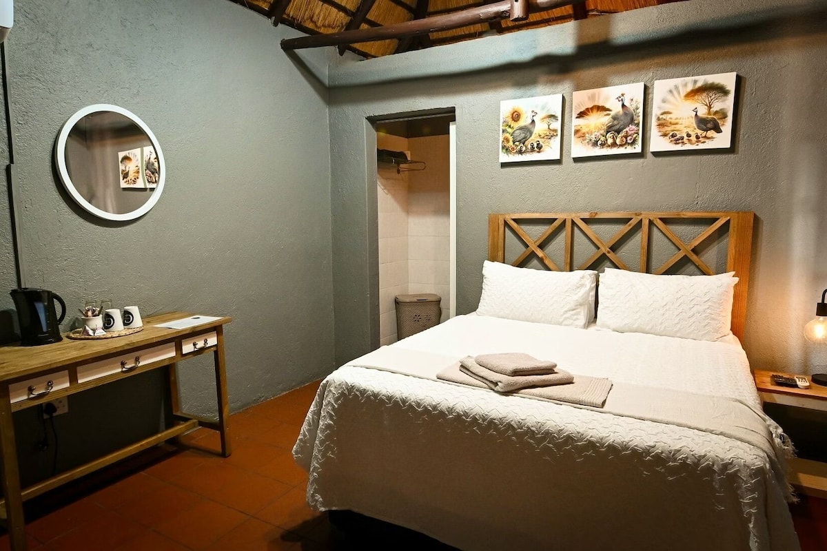 Quarto com cama, escrivaninha de madeira, espelho redondo e três quadros pendurados na parede.