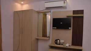 Quarto de hotel com guarda-roupa, espelho, televisão, mesa pequena com chaleira e chávenas, e um cesto de lixo.