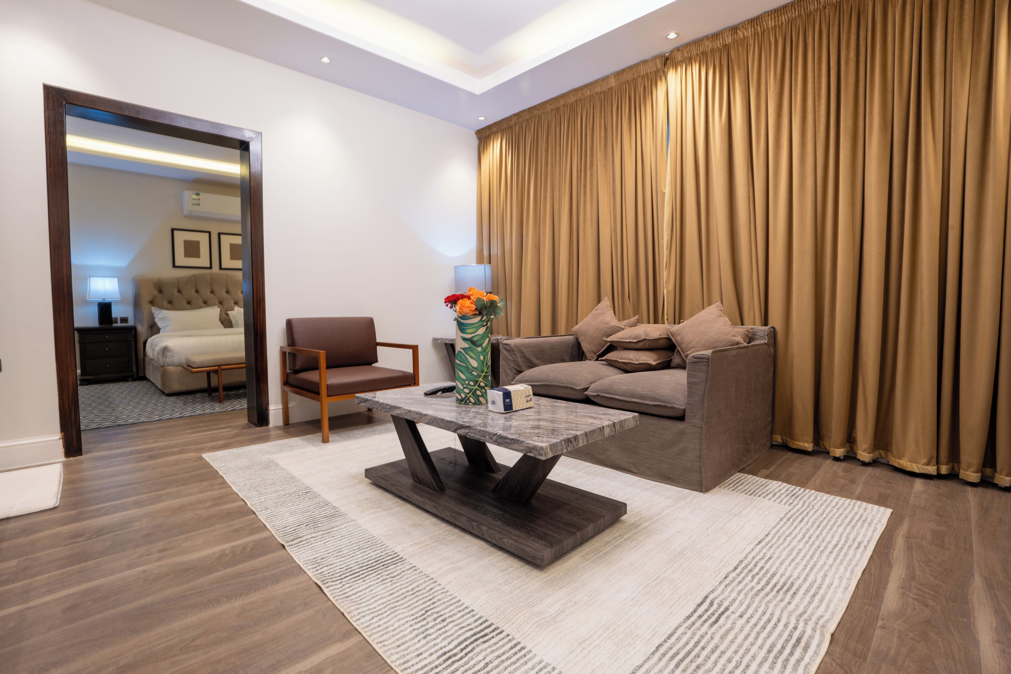 Uma sala de estar moderna com um sofá, poltrona e mesa de centro. Há uma janela grande com cortinas, um espelho e uma porta que dá para outro cômodo.