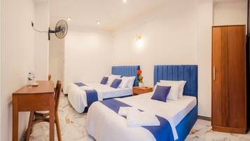 Quarto de hotel com duas camas, uma escrivaninha de madeira, uma cadeira, um guarda-roupa e um ventilador fixado na parede.
