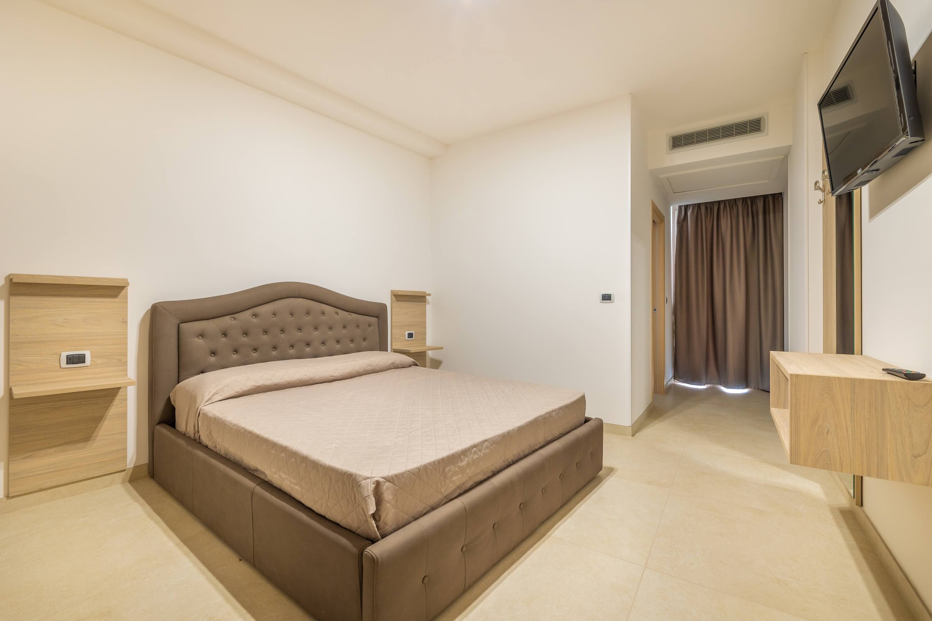 Quarto moderno com cama grande, mesas de cabeceira, televisão e escrivaninha embutida.