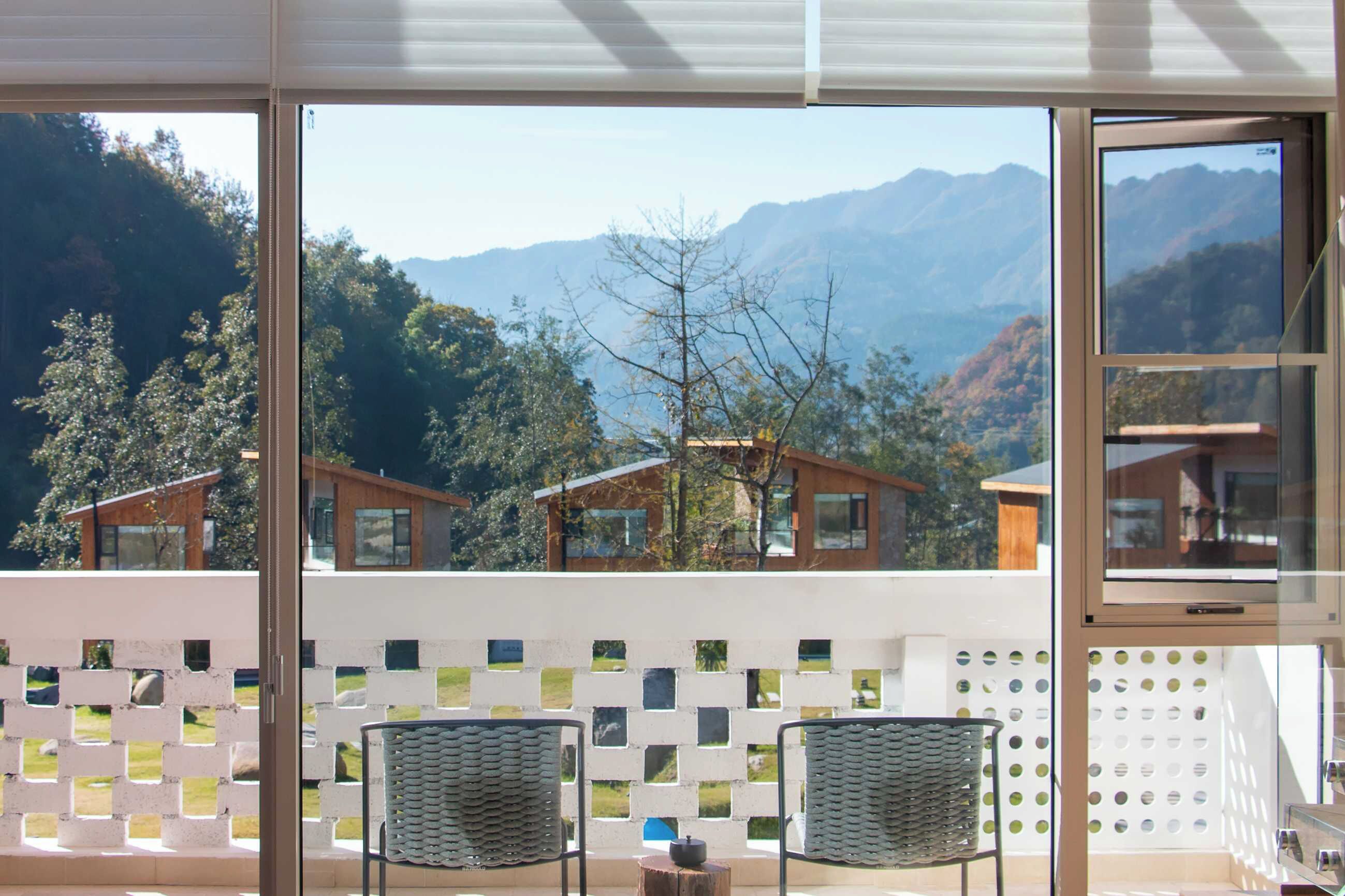Uma varanda com duas cadeiras, uma mesa pequena e vista para montanhas e casas.