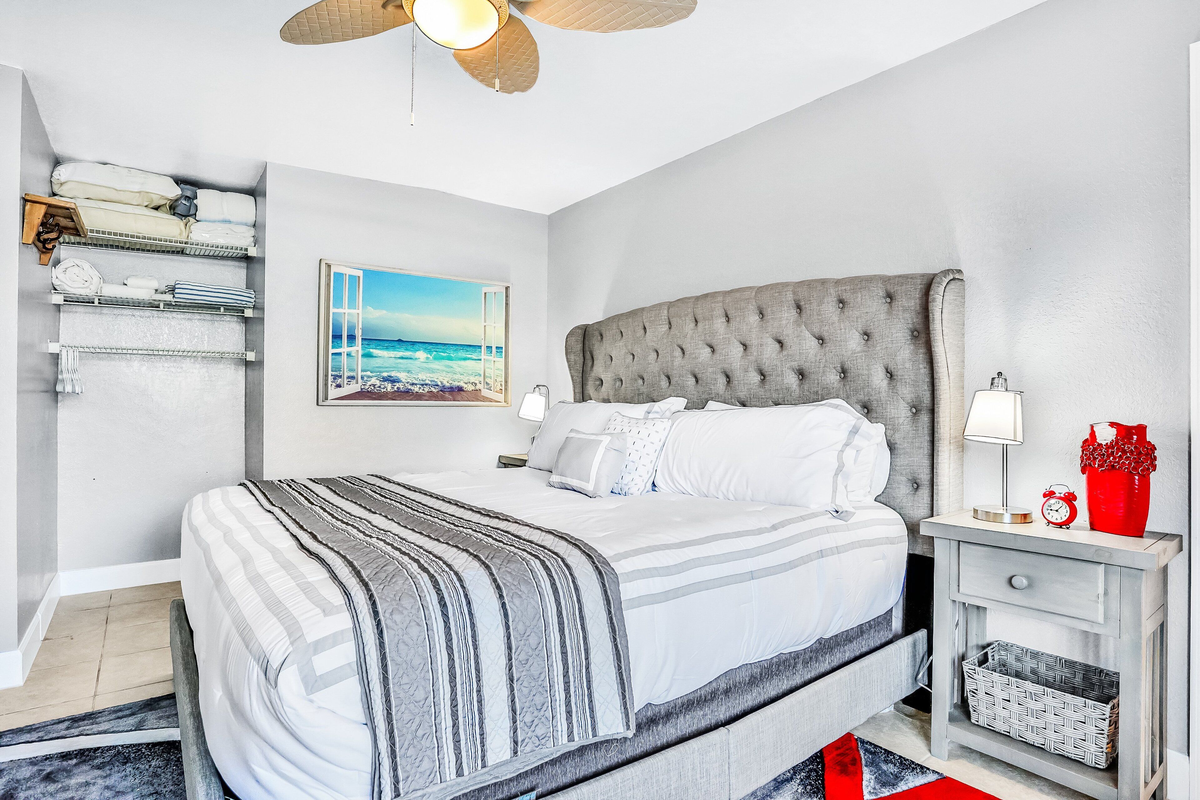 Quarto com cama grande, mesa de cabeceira, prateleira na parede com toalhas e quadro com vista para o mar.