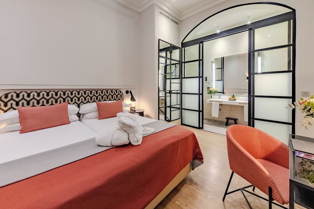 Quarto de hotel moderno com uma cama grande, uma poltrona vermelha estofada e um painel de vidro separando o quarto do banheiro.