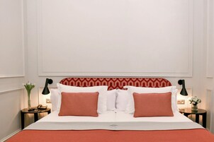 Cama bem arrumada com roupa de cama branca e vermelha, duas mesas de cabeceira com abajures e uma cabeceira estampada.