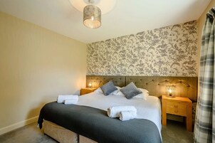 Cama bem arrumada com colcha escura e duas almofadas, com mesas de cabeceira de madeira e luminárias.