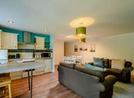 Uma cozinha moderna com armários brancos e bancada cinza, uma sala de jantar com mesa e cadeiras brancas, e uma sala de estar com sofá cinza e almofadas azuis.