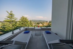 Uma varanda com duas cadeiras de descanso brancas, uma mesa pequena e vista para uma paisagem verde com árvores e edifícios ao longe.