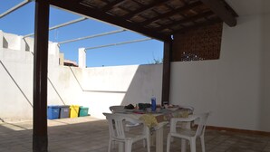 Um pátio externo com mesa e cadeiras, piso de azulejos e área coberta com vista para um edifício e céu.