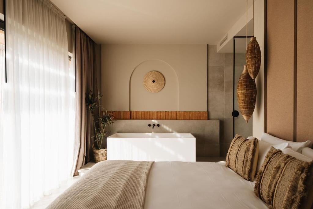Quarto de hotel moderno com uma cama grande, uma banheira integrada e uma janela ampla com cortinas.