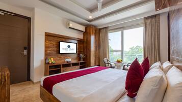 Quarto de hotel com cama, televisão fixada na parede, escrivaninha com cadeira e janela com cortinas.