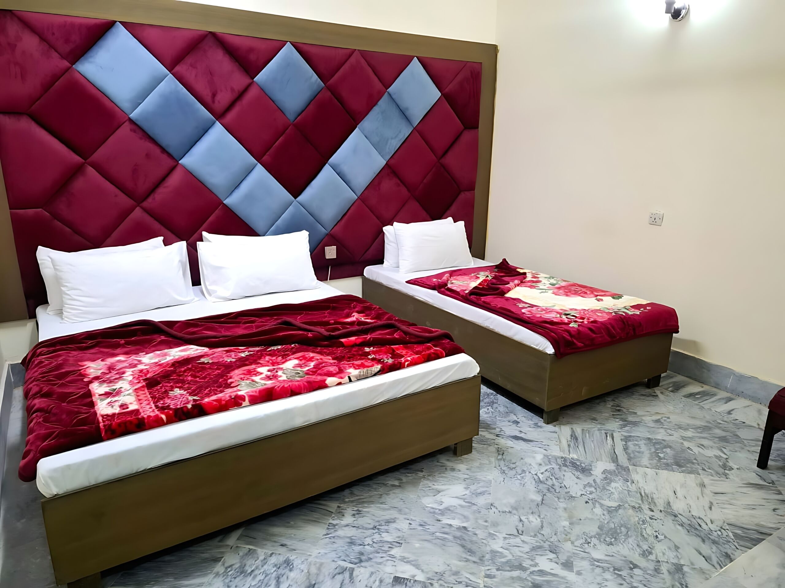 Quarto de hotel com duas camas, cada uma com colcha floral vermelha e almofadas brancas. A cabeceira é vermelha com padrão de losangos azuis.