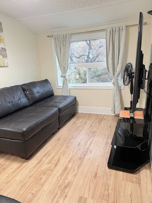 Uma sala de estar com sofá de couro preto, piso de madeira, janela com cortinas e um rack preto para TV com controle remoto.