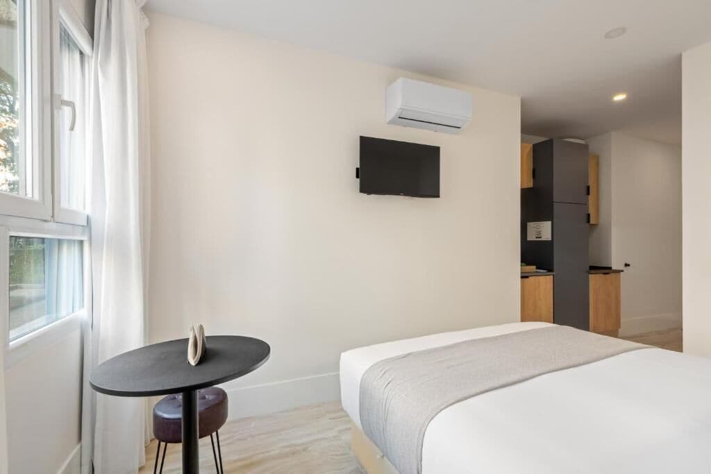 Quarto de hotel moderno com uma cama, uma mesa redonda pequena, uma TV de tela plana na parede e um aparelho de ar condicionado.