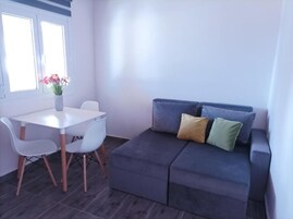 Quarto pequeno e moderno com um sofá cinzento, uma mesa branca e duas cadeiras. Há uma janela com molduras brancas e um vaso com flores sobre a mesa.