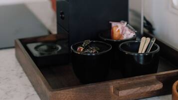 Máquina de café sobre bandeja de madeira, com duas chávenas pretas e um pequeno recipiente, com uma parede branca e uma planta verde ao fundo.