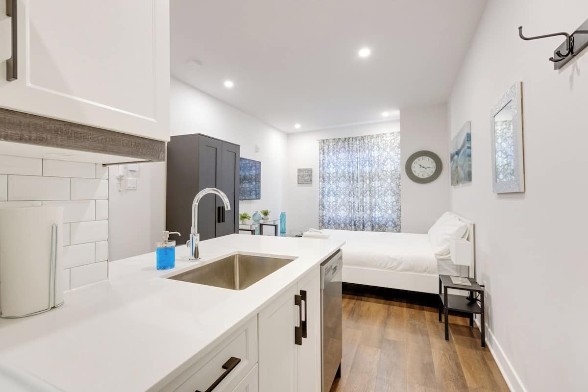 Quarto de hotel pequeno com cozinha compacta, uma cama, mesa de cabeceira e pequena escrivaninha.