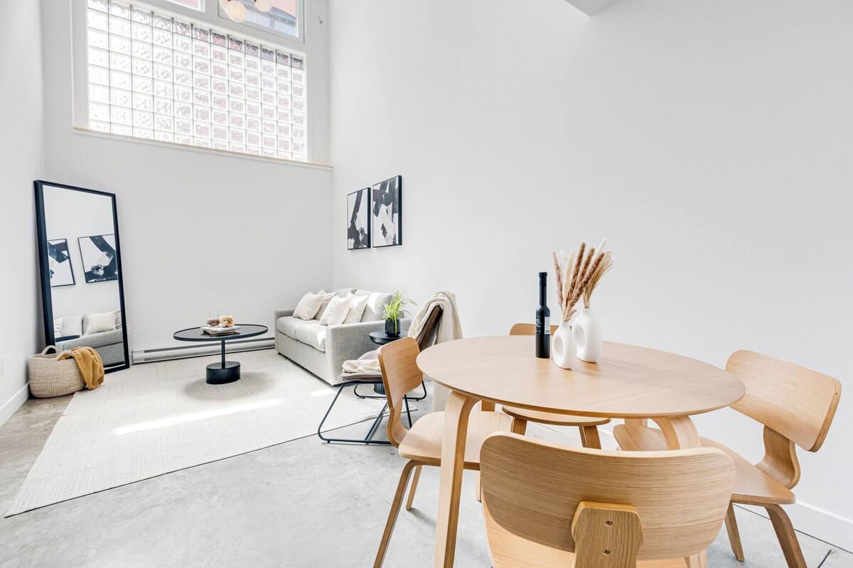 Uma sala de estar moderna com uma mesa de madeira redonda, sofá bege e uma grande janela com persianas.