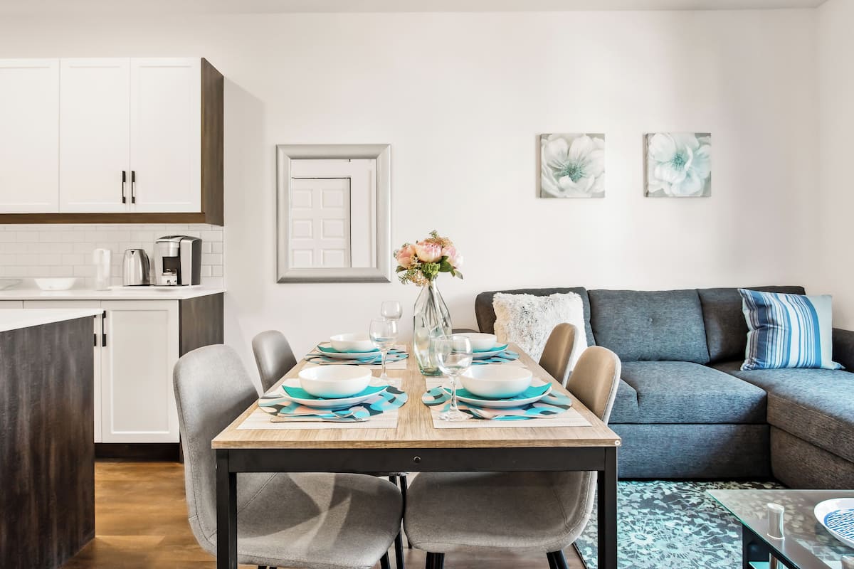 Cozinha moderna com armários brancos, mesa de jantar de madeira posta com jogos americanos azul-turquesa, e sofá cinza na sala de estar.