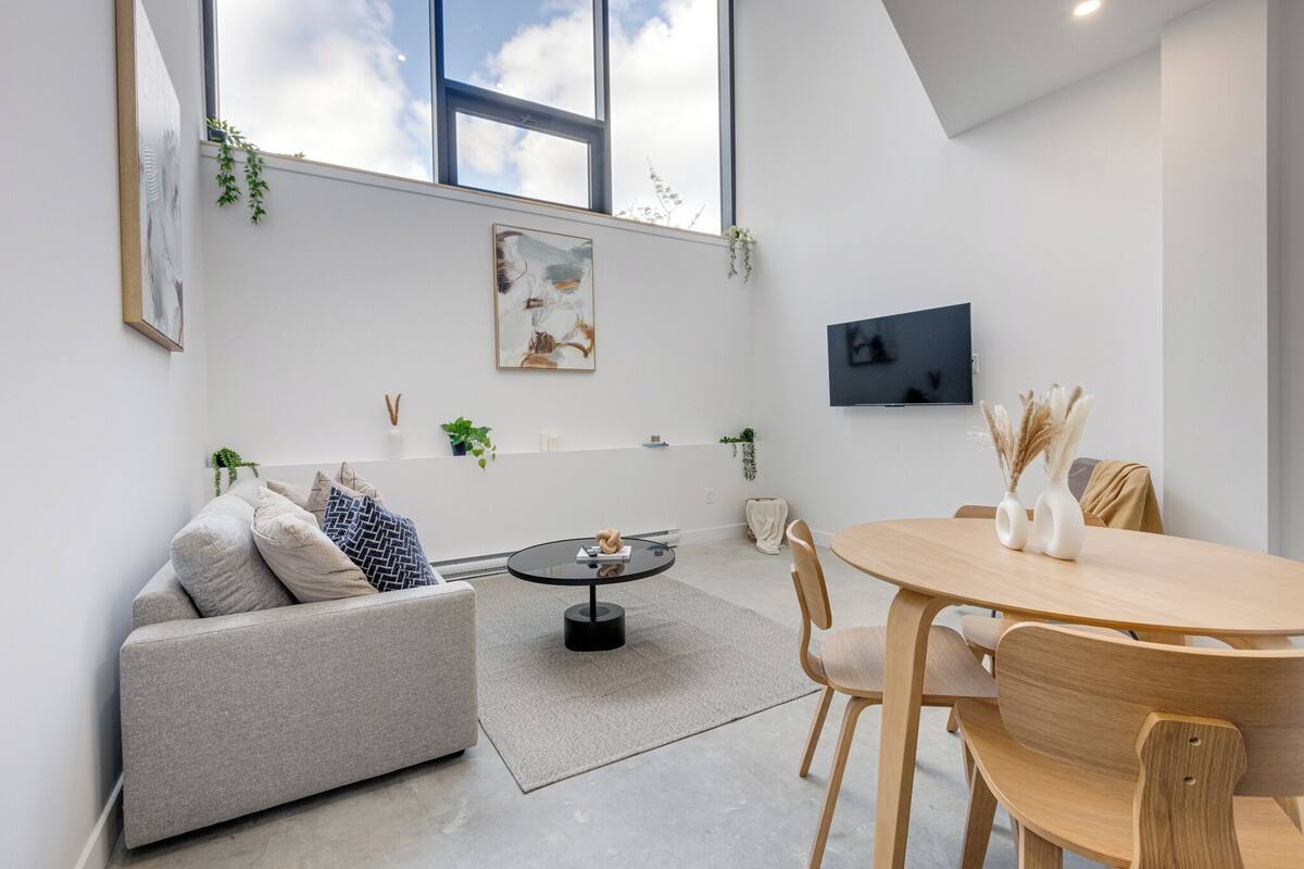 Uma sala de estar moderna com um sofá cinzento, uma mesa de centro redonda e uma mesa de jantar de madeira com cadeiras. Há plantas nas paredes e uma televisão de tela plana montada acima da mesa de centro.