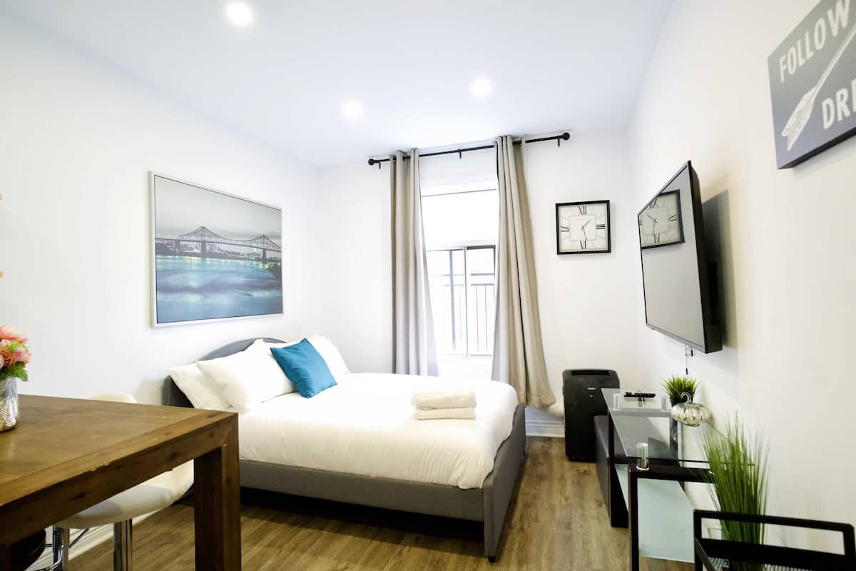 Quarto de hotel moderno com cama, escrivaninha, cadeira, televisão e janela com cortinas.