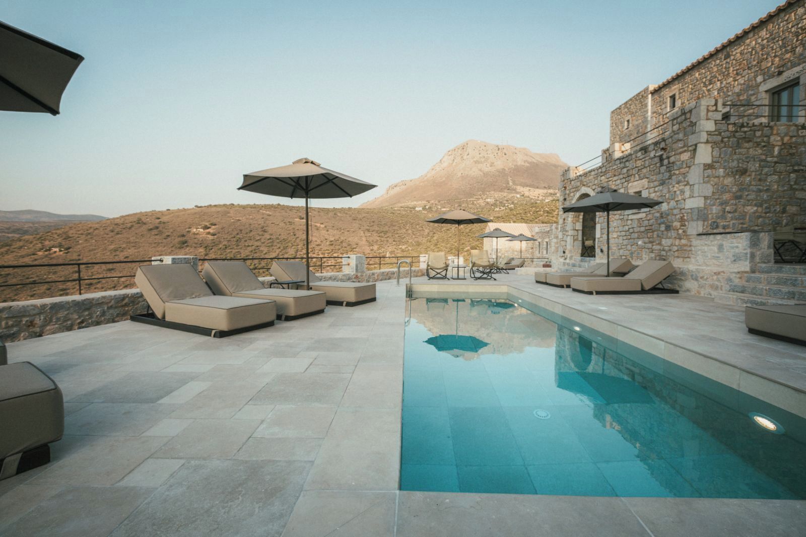 Área de piscina com espreguiçadeiras e guarda-sóis, paredes de pedra e montanhas ao fundo.