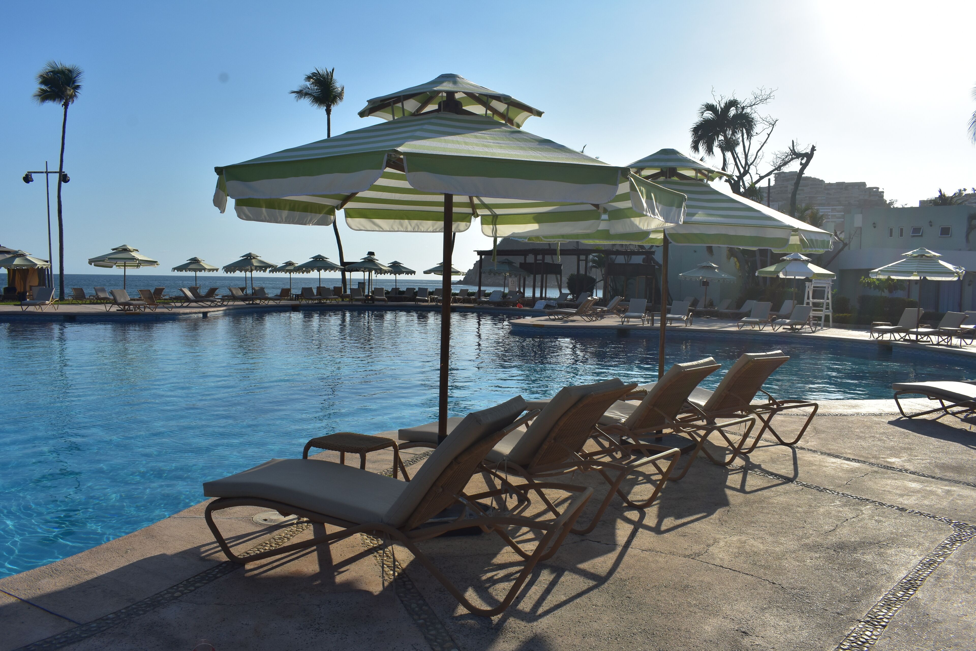 Uma área na piscina com espreguiçadeiras e guarda-sóis listrados, vista desimpedida do oceano e um edifício ao fundo.