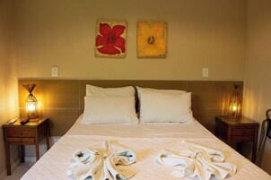 Quarto de hotel com uma cama grande, duas mesas de cabeceira, uma luminária e duas pinturas na parede.