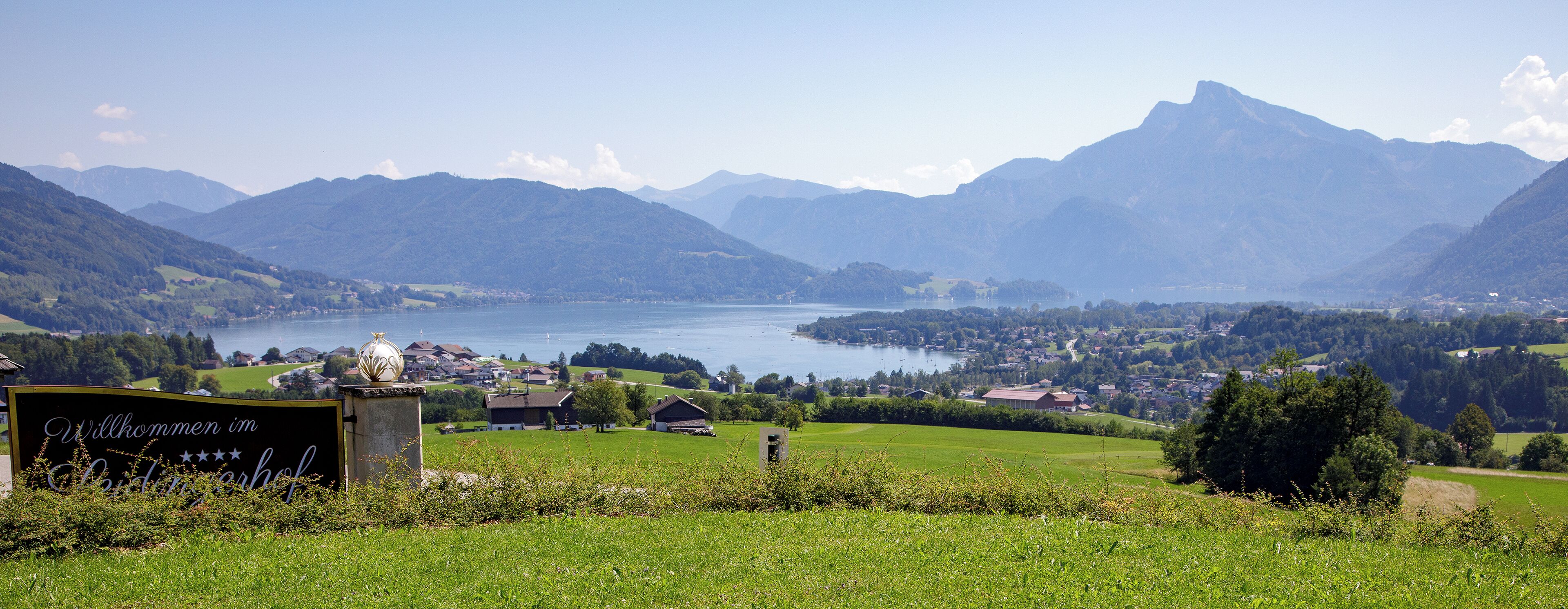 Uma vista panorâmica de um lago montanhoso, com uma aldeia em primeiro plano e montanhas ao fundo.