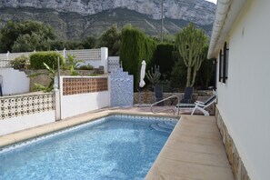 Piscina com borda azulejada, rodeada por um muro branco e um muro de pedra. Há espreguiçadeiras e uma mesa perto da piscina.